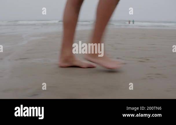Teenager walking barefoot on sandy shores, enjoying a cloudy day by the ocean, creating joyful ...