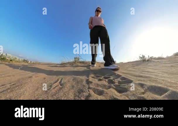 A worm's-eye view of a woman walking on sandy ground with sparse dry ...