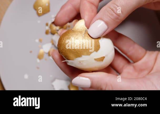 Detailed shot of hands peeling a golden-painted hard-boiled Easter egg, with shiny fragments ...