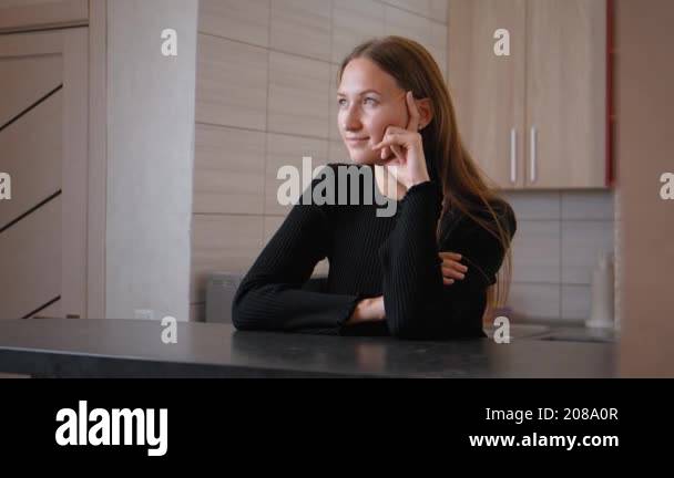 Young woman leans on her kitchen counter, resting her chin on her hand ...