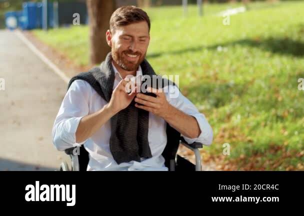 Smiling young disabled man use phone on a wheelchair in a park at ...