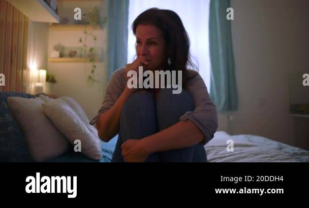 Woman sitting on bed, knees pulled close, tears in her eyes, overwhelmed with sadness, themes of ...
