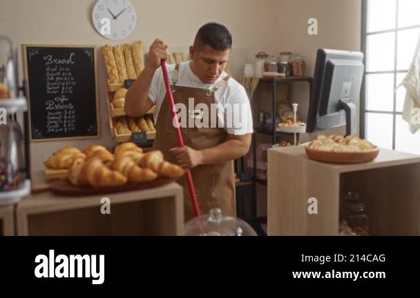 Young man in an apron cleans a bakery, leaning tiredly on a mop ...
