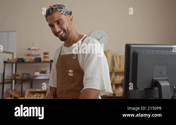 Young man working in a bakery shop, wearing a brown apron and smiling ...