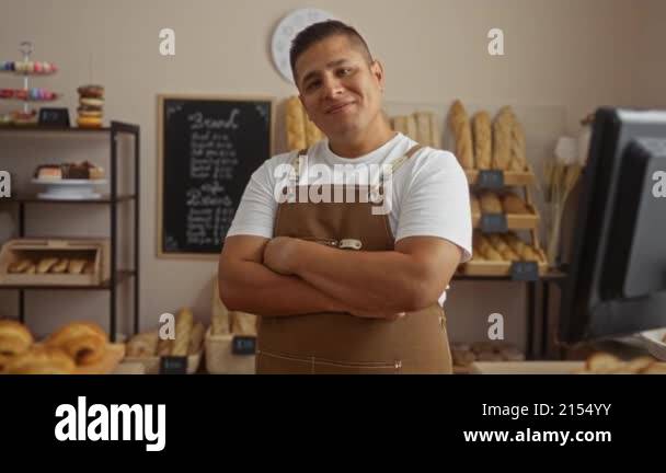 Young man wearing apron giving thumbs up while standing in bakery with crossed arms against ...