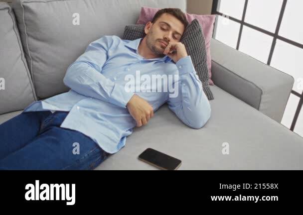 A handsome young hispanic man with a beard wearing a blue shirt and jeans is seen indoors in his ...