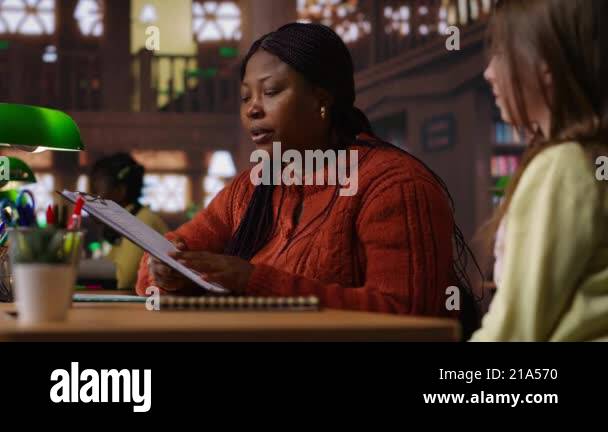 Black tutor mentoring schoolgirl in a school library study desk ...
