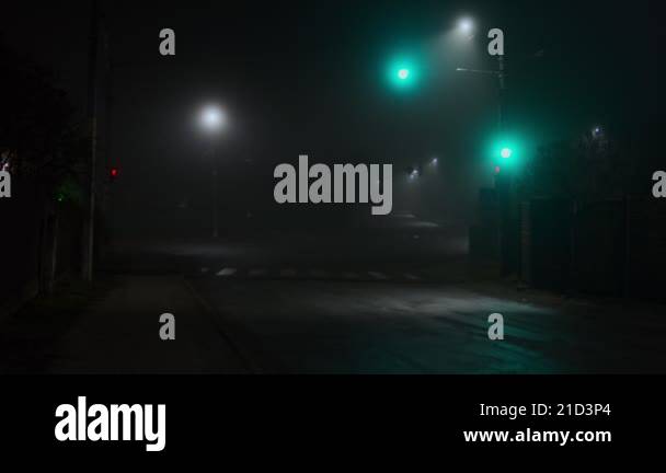 A deserted street at night with fog, lit by green traffic lights and ...