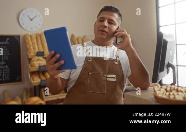 Young man in brown apron using blue tablet and talking on phone in ...