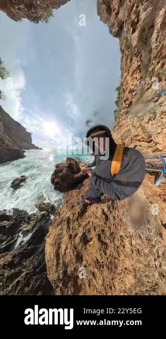 Vertical video. A distorted wide-angle shot of a man standing on rocky cliffs near the ocean ...