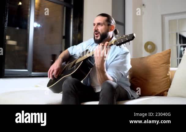bearded man in shirt plays black acoustic guitar and singing at home ...