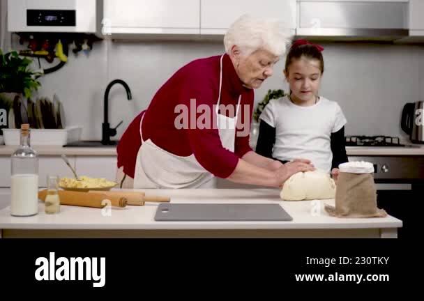 Loving grandmother teaches sweet granddaughter to knead yeast dough with hands on table. Little ...