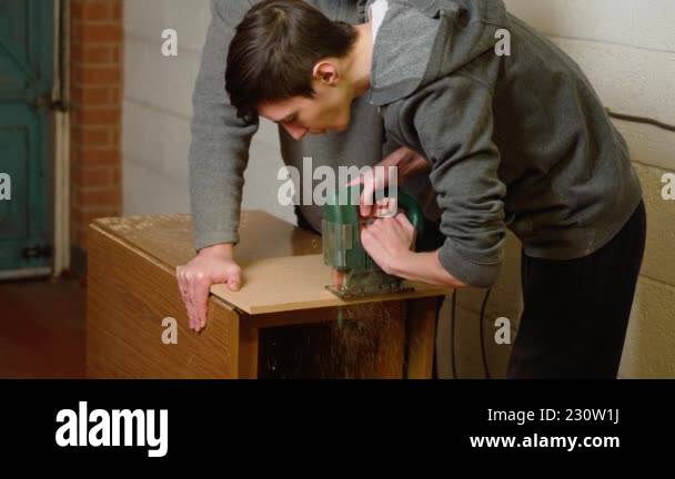 Father with son cutting wooden board using jigsaw in home workshop. Dad assisting while boy ...