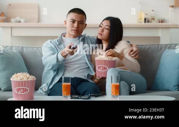 A couple enjoys a cozy evening at home, sitting on a couch with popcorn ...