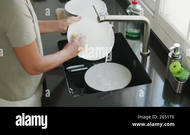 Woman washing and rinsing dishes by hand under a stream of water in the ...