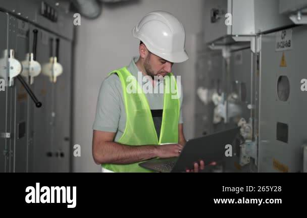 Serious electrical engineer using laptop near electrical switchboard. Focused man working with ...
