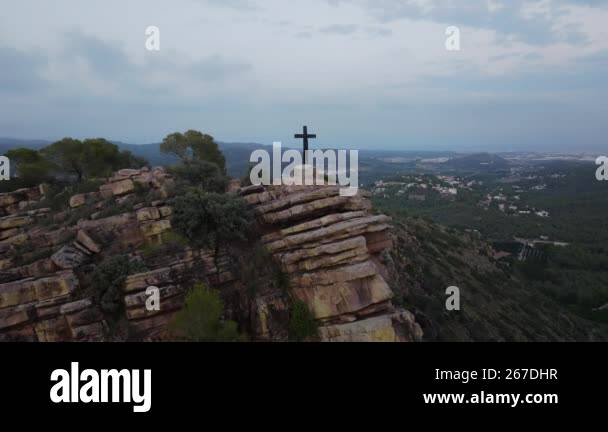 A black cross stands atop a rocky cliff, surrounded by lush greenery ...