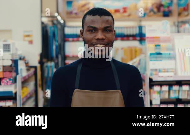 Smiling male store clerk wearing an apron, standing in a well-organized ...