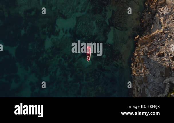 A red boat floats in dark, clear waters near a rocky coastline. The ...