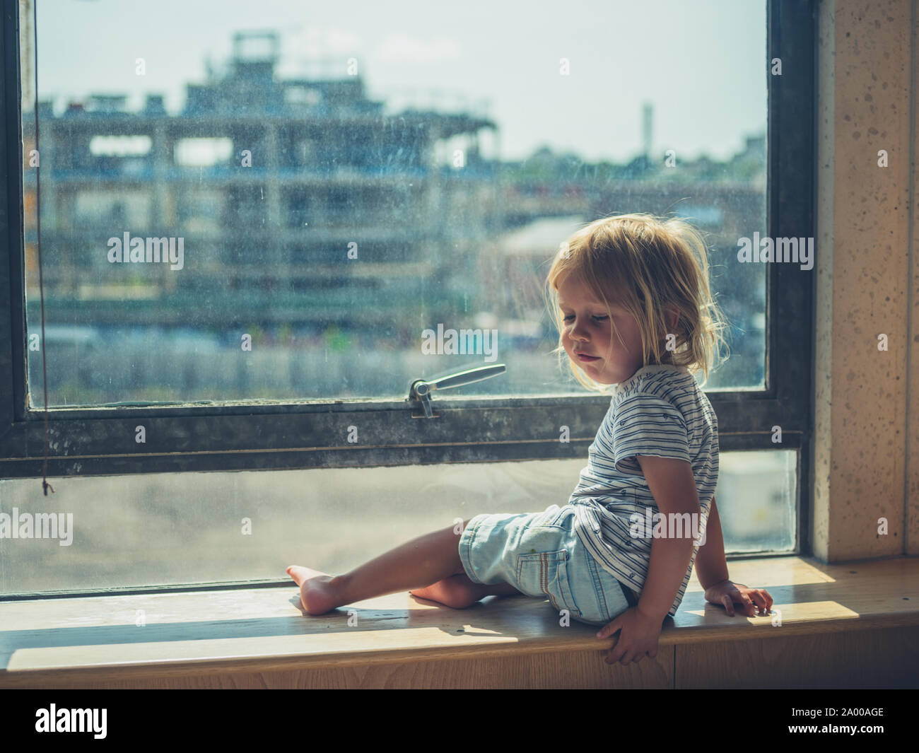 A little toddler is sitting by the window in a city apartment looking ...