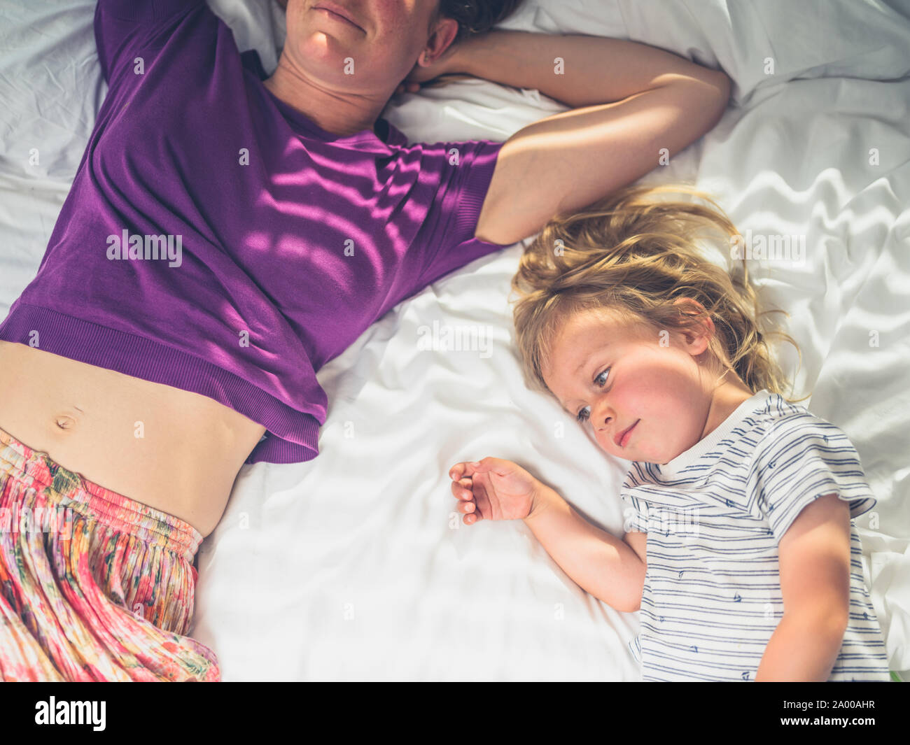 A little toddler is relaxing on a bed with his mother in the sunshine ...