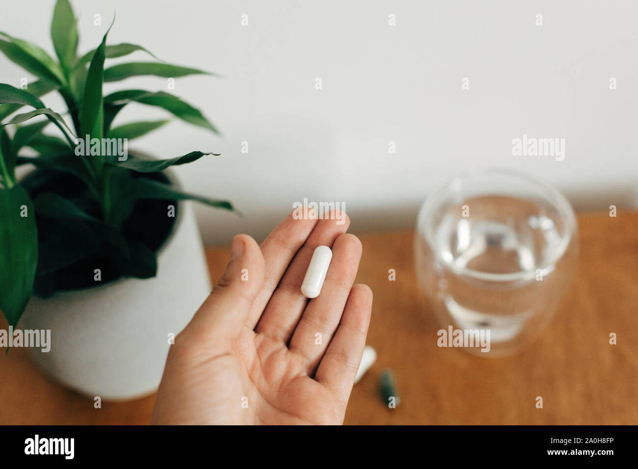 Hand holding magnesium capsule above glass of water on wooden table. White pill. Dietary ...