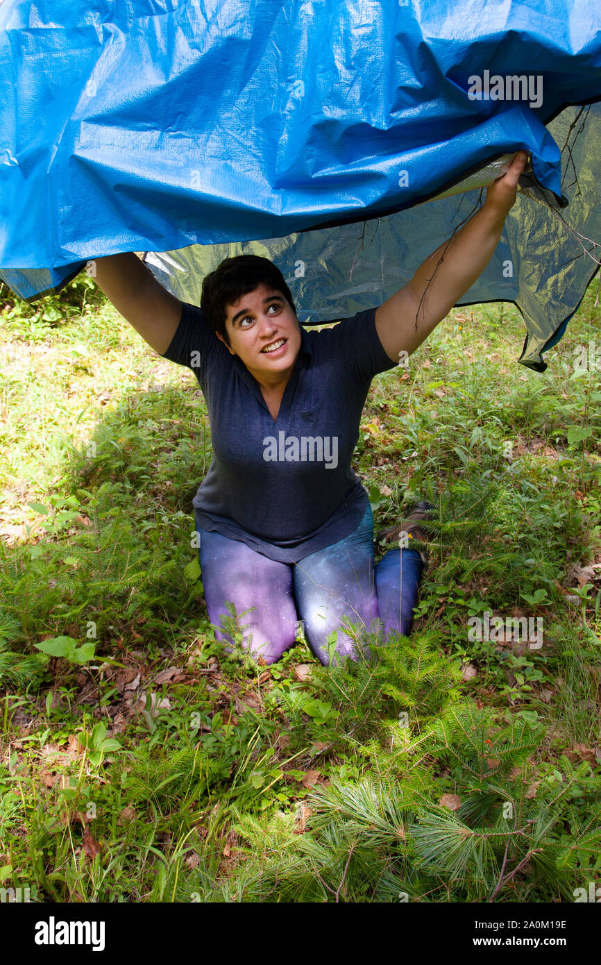 person takes shelter under a blue tarp in the forest while camping ...