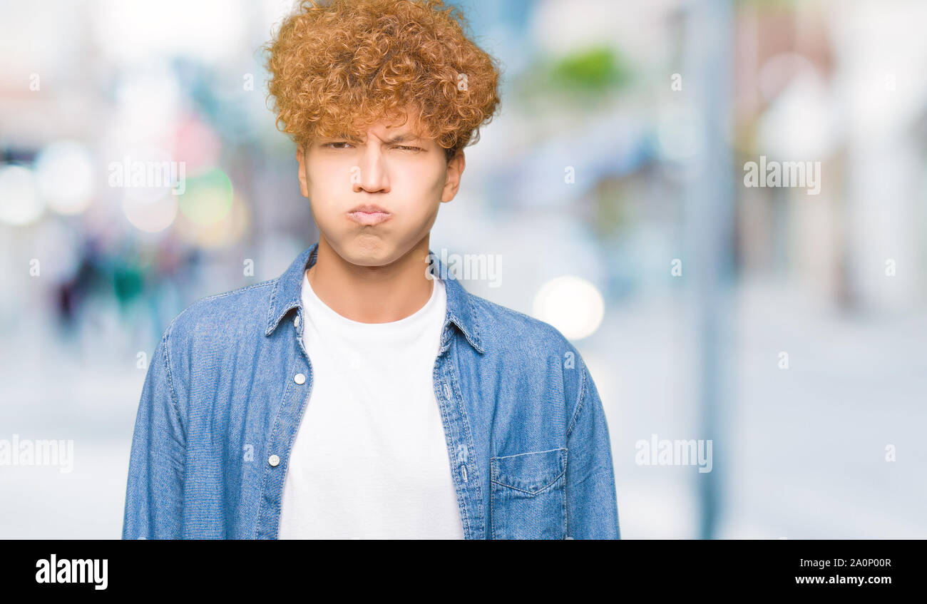 Young handsome man with afro hair wearing denim jacket puffing cheeks ...