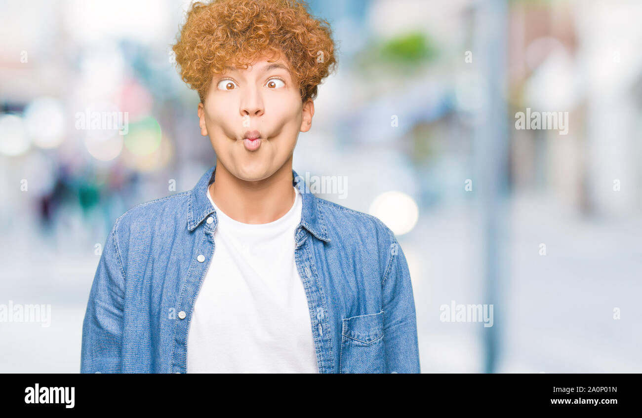 Young handsome man with afro hair wearing denim jacket making fish face ...