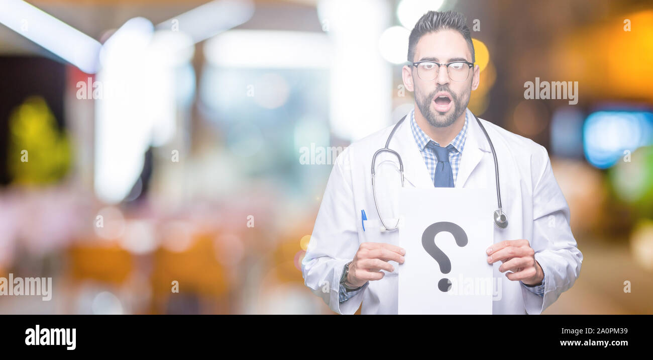 Handsome young doctor man holding paper with question mark over ...