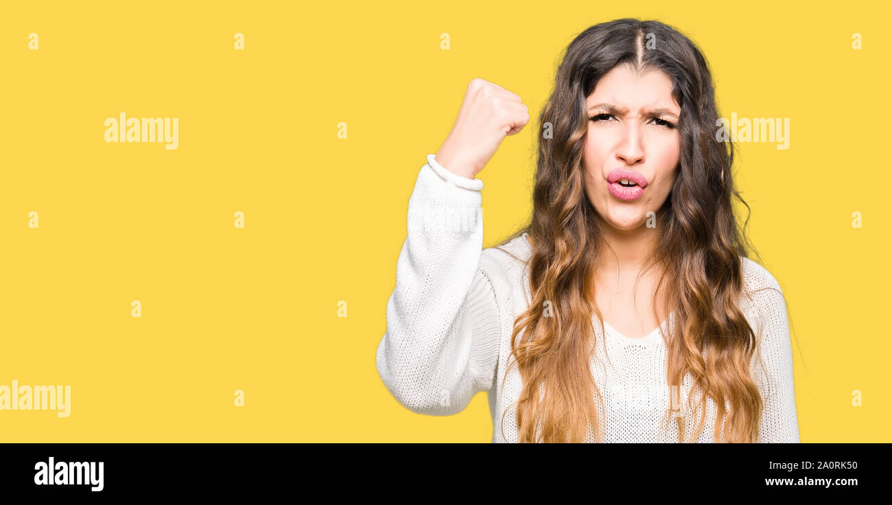 Young beautiful woman wearing white sweater angry and mad raising fist ...
