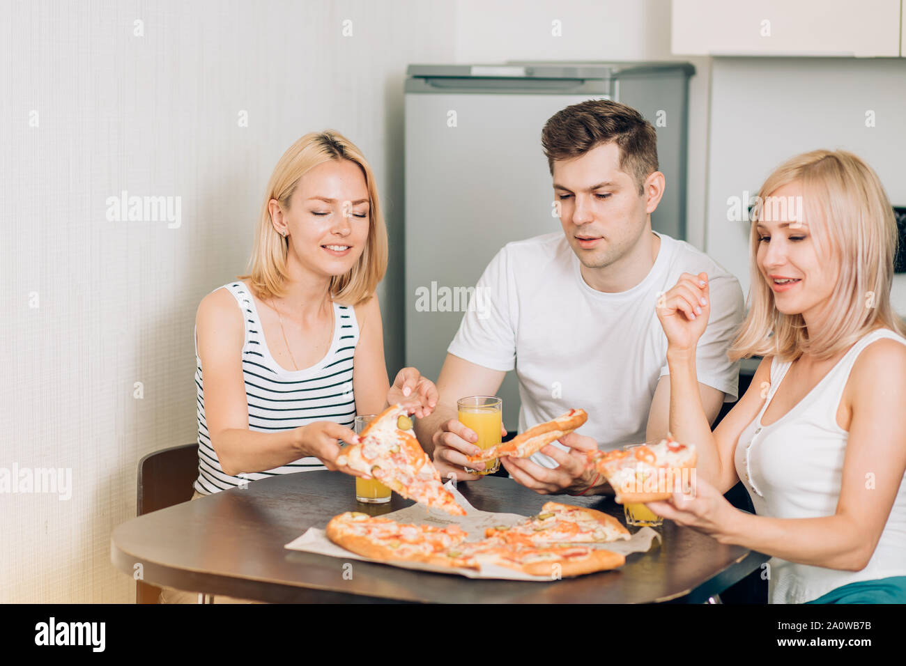 Three caucasian happy students eating pizza, drinking orange juice in ...