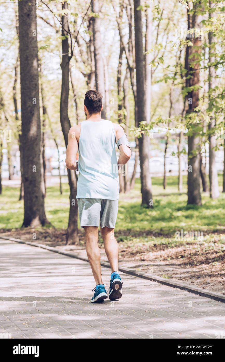 back view of young man in sportswear jogging along walkway in park ...