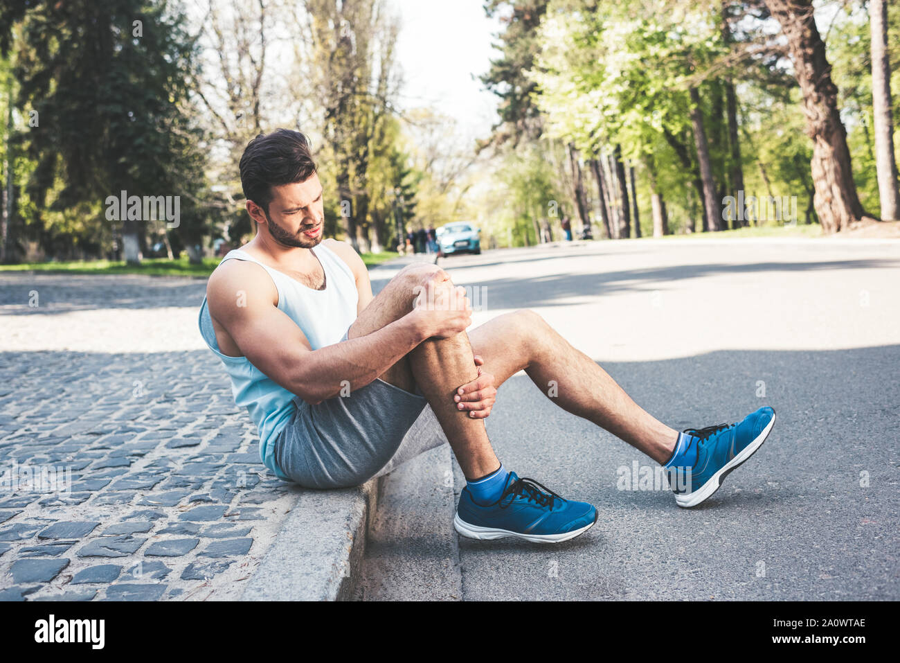 young sportsman sitting on pavement border and touching injured leg ...