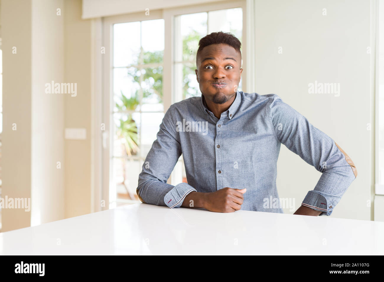 Handsome african american man on white table puffing cheeks with funny ...