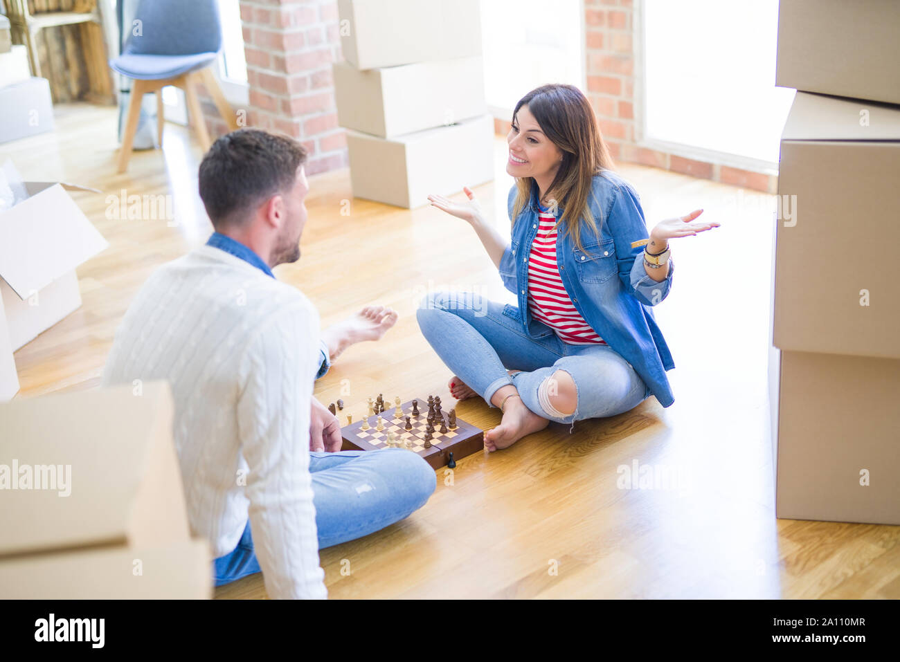 Young beautiful couple playing chess at new home around cardboard boxes ...