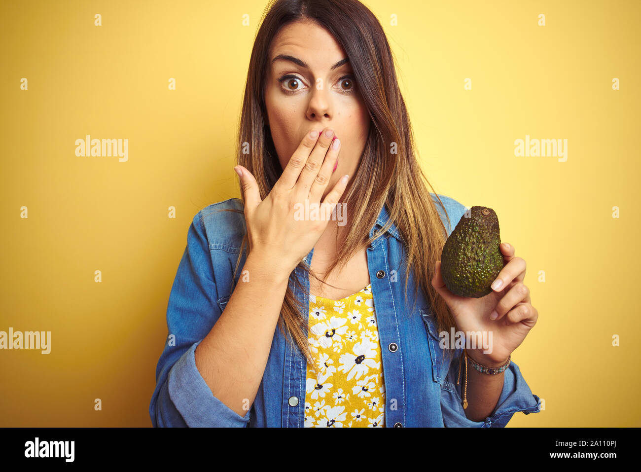 Young beautiful woman eating healthy avocado over yellow background ...