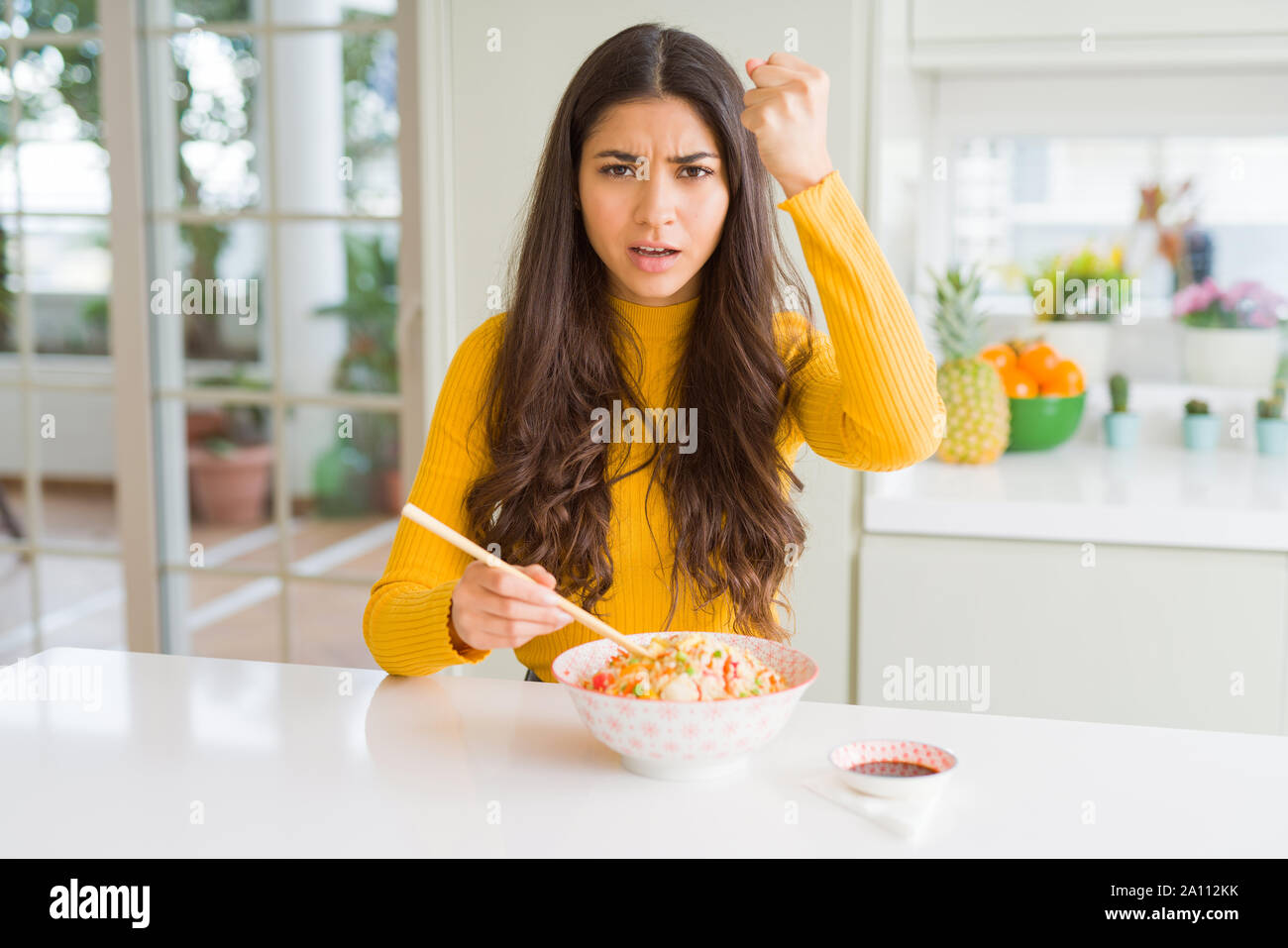 Young woman eating a bowl of Asian rice using chopsticks annoyed and ...