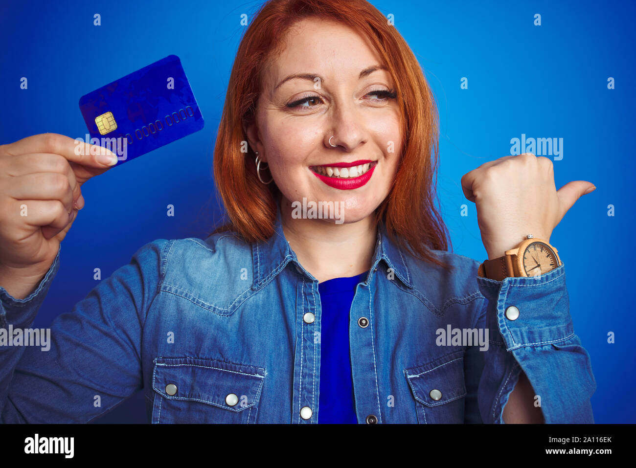 Young beautiful redhead woman holding credit card over blue isolated ...
