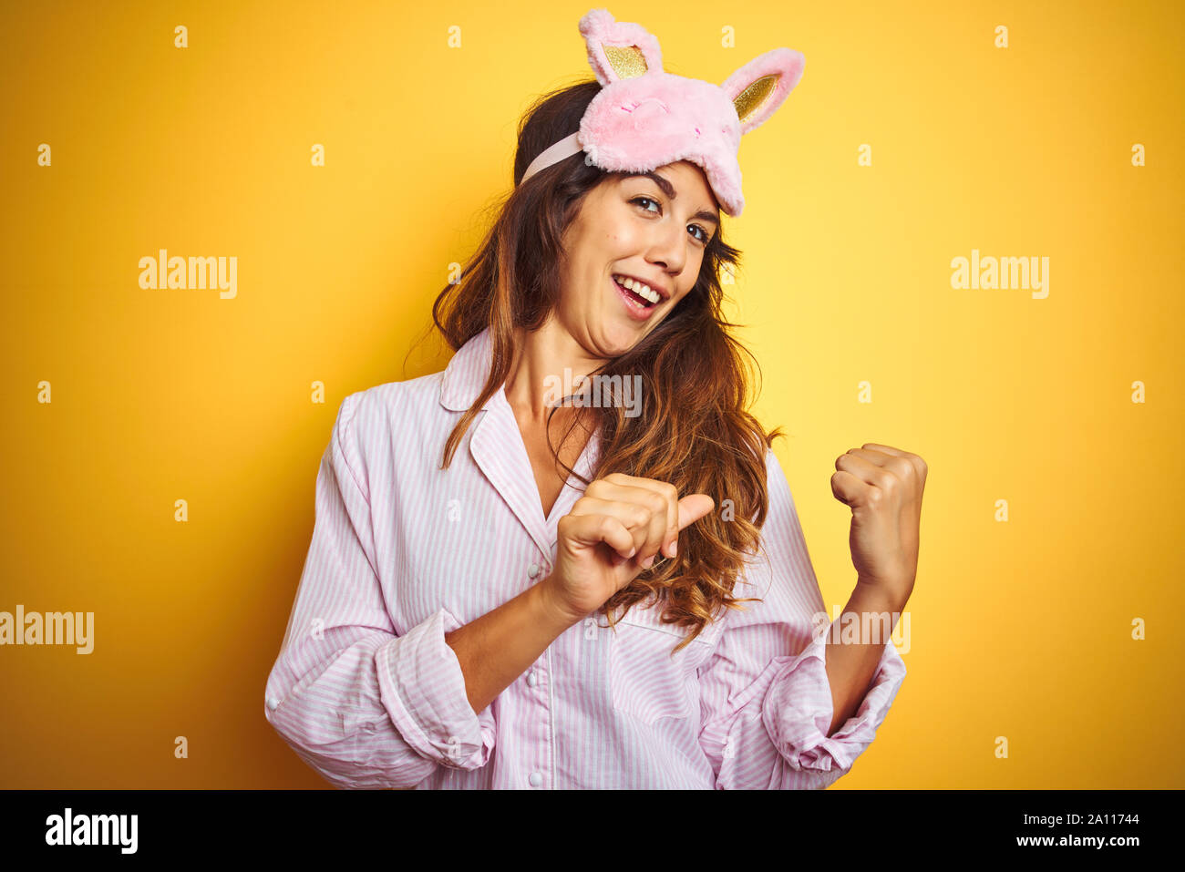 Young woman wearing pajama and sleep mask standing over yellow isolated ...