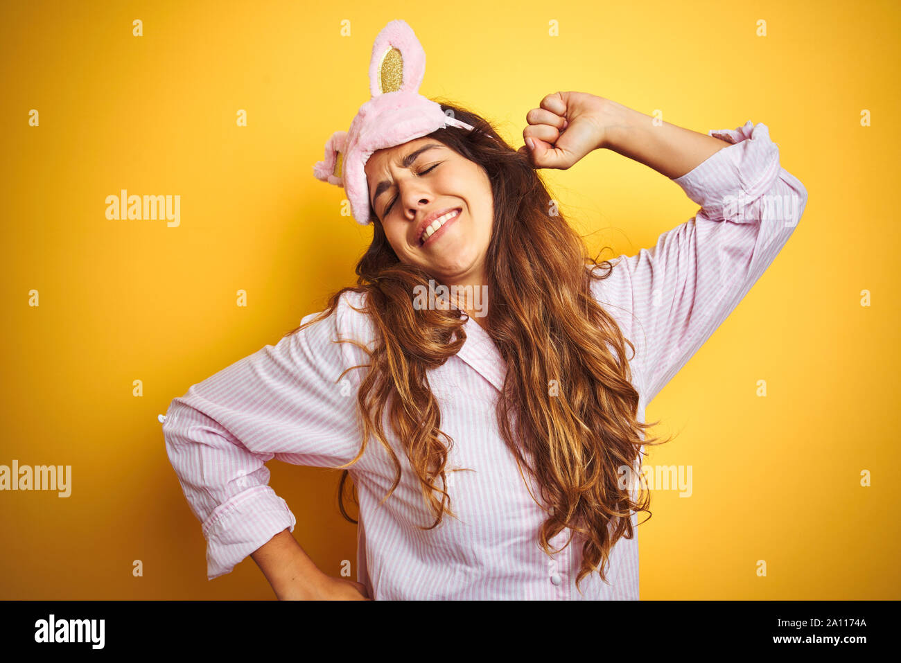 Young woman wearing pajama and sleep mask standing over yellow isolated ...