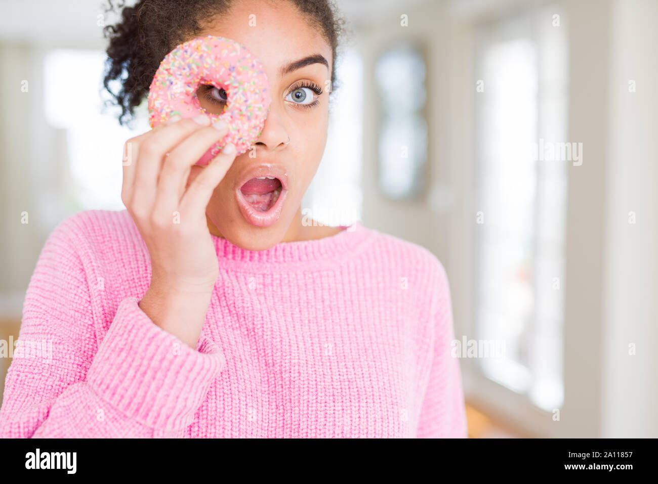 Young african american girl eating sweet pink donut scared in shock ...