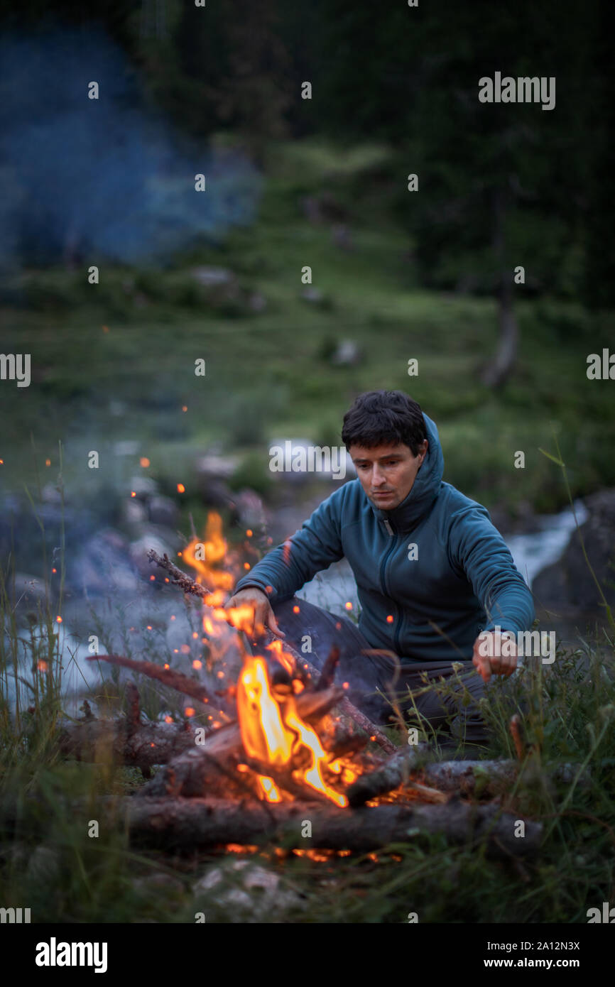 Young man making fire while camping outdoors, in an alpine wilderness ...