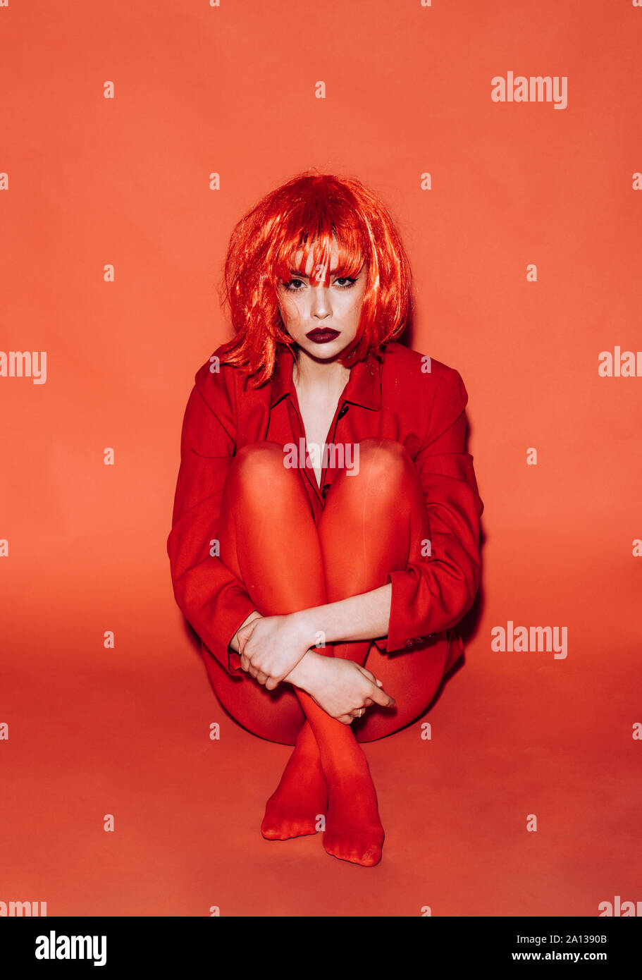 Serious young female in red wig, tights and jacket sitting on floor ...