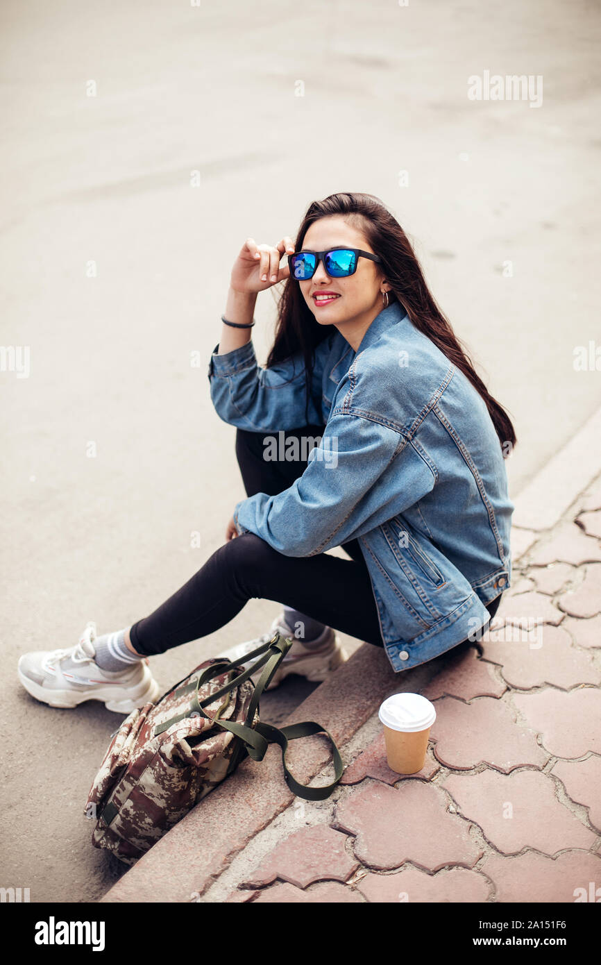 Young girl student is sitting on the pavement. Wear jeans and drink ...