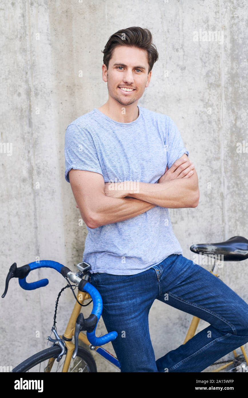 Portrait of smiling man with racing cycle standing in front of concrete ...