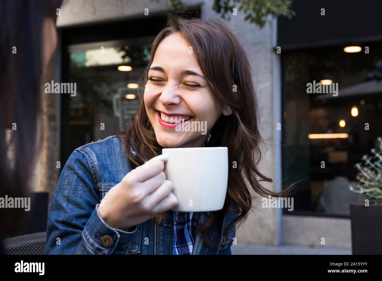 Happy young woman laughing with eyes closed and holding coffee cup in outside cafe in Madrid ...