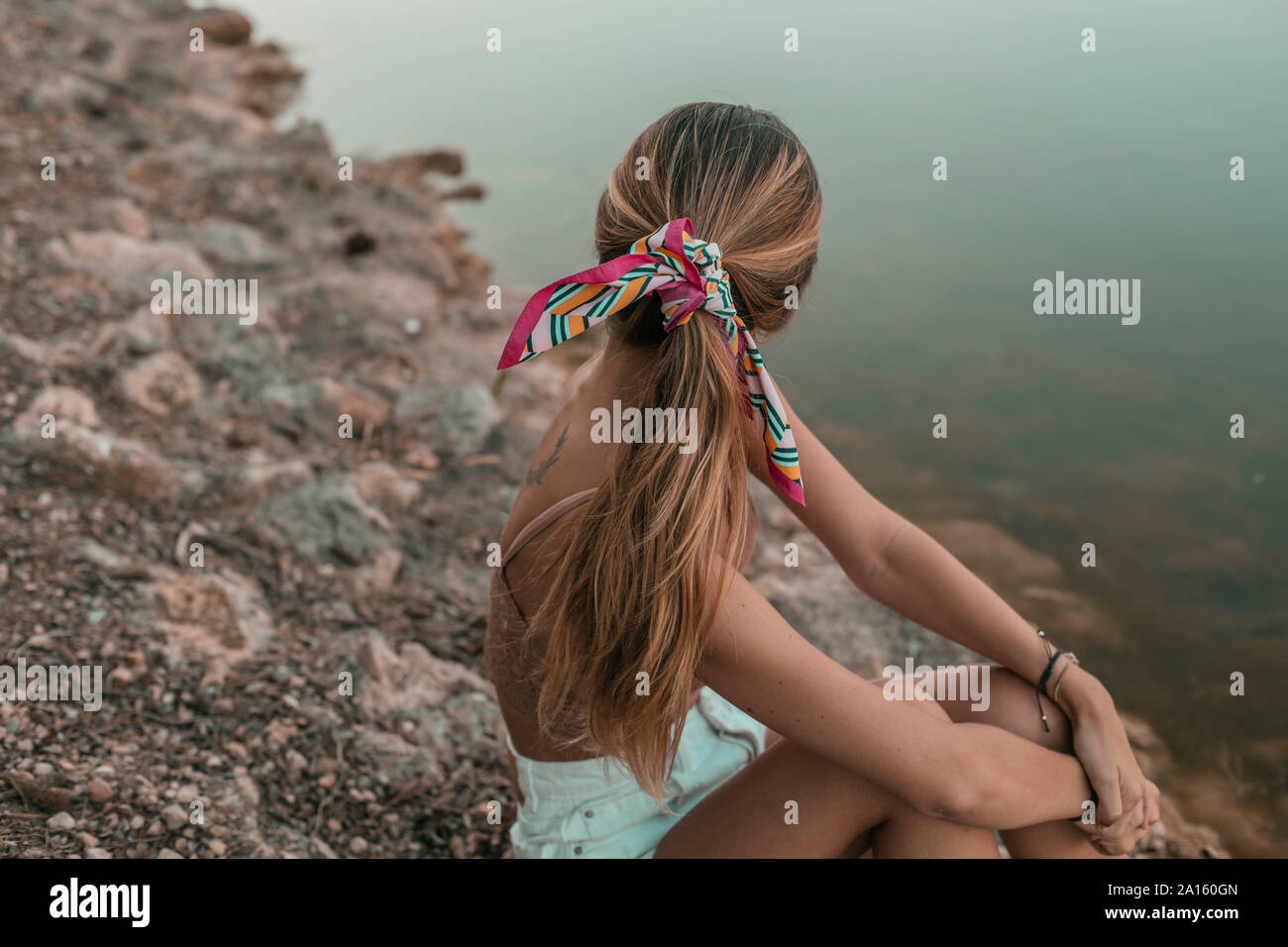 Rear view of young blond woman sitting at a lakeside, hair with cloth ...