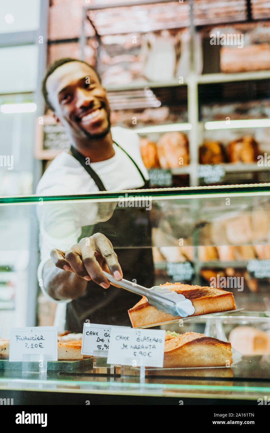 Smiling man working in a bakery taking a piece of cake with a tongs ...