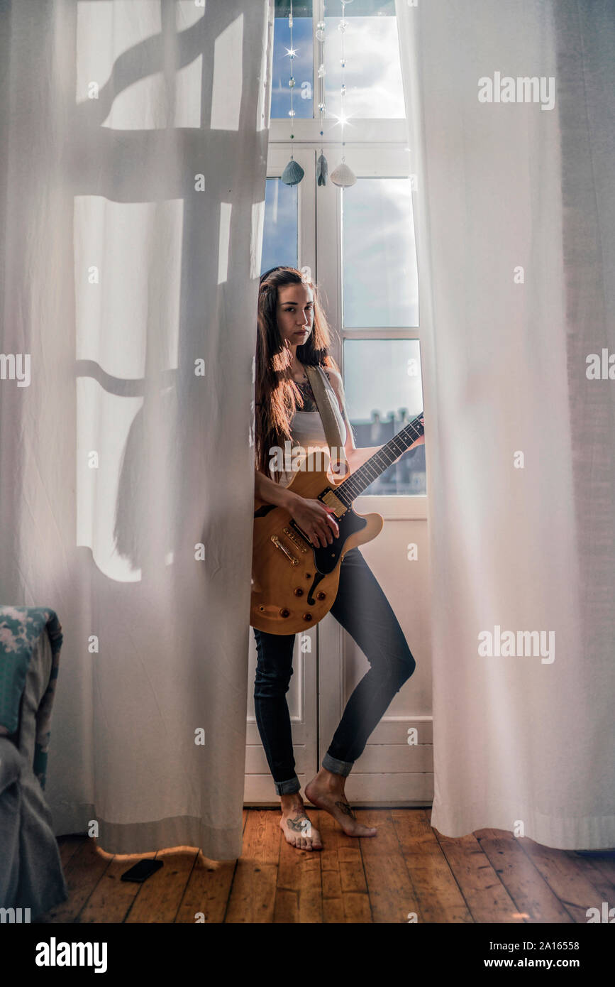 Serious young woman standing at the window at home with a guitar Stock ...